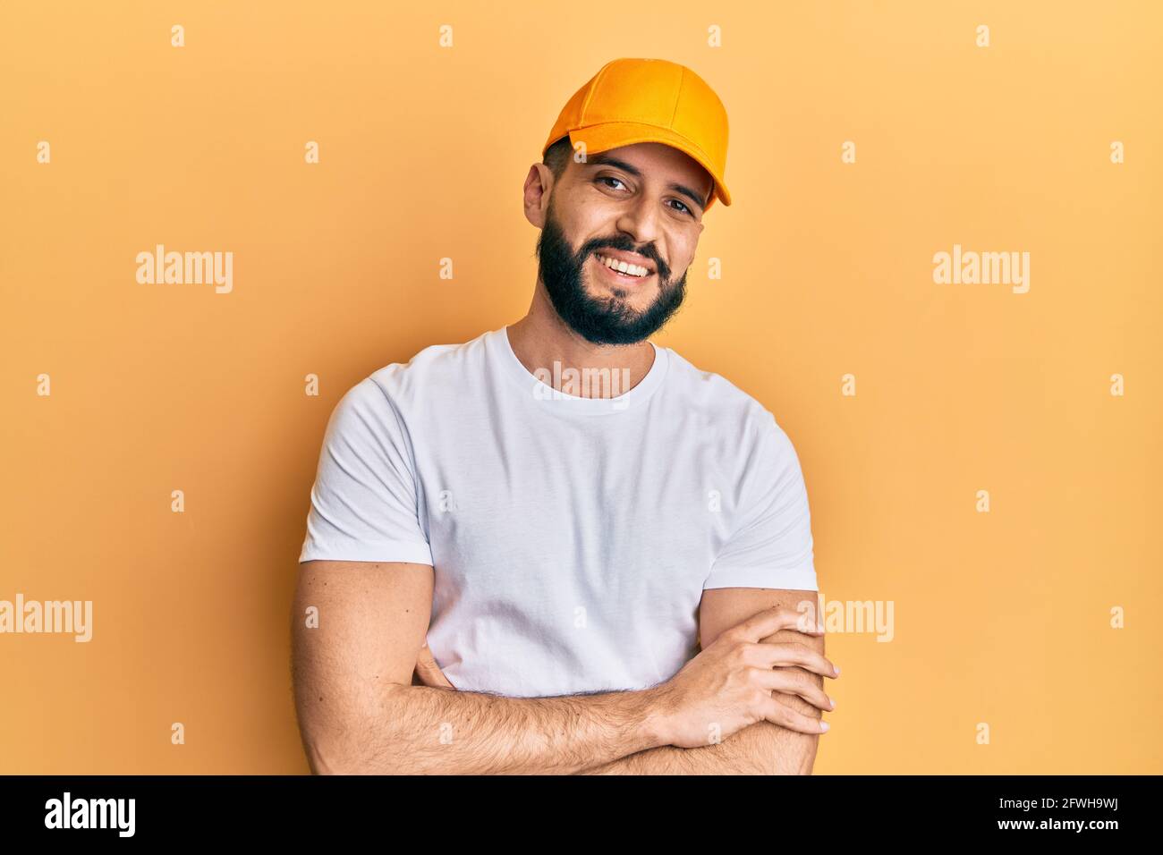 Young man with beard wearing yellow cap happy face smiling with crossed ...