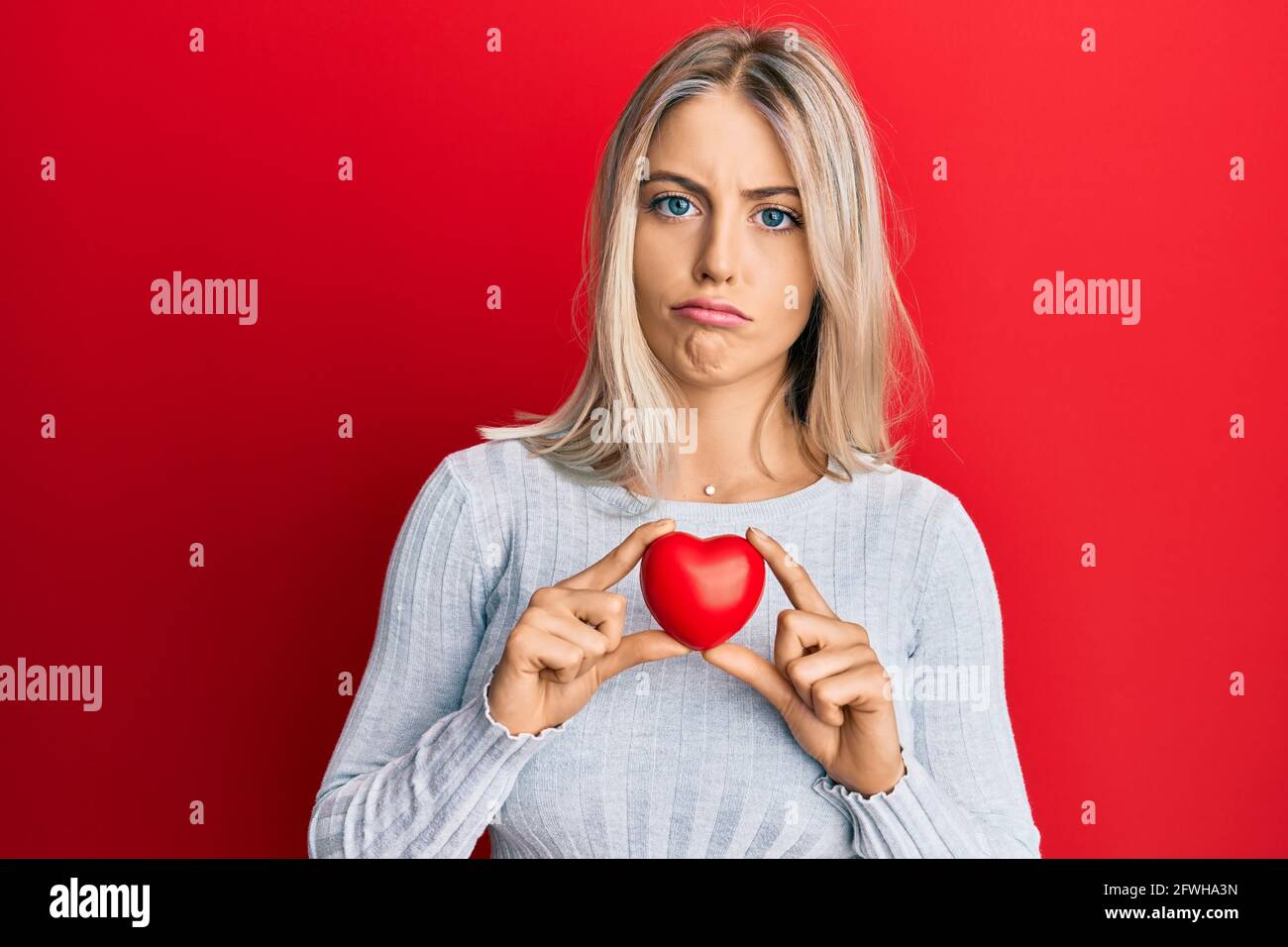 Beautiful blonde woman holding heart depressed and worry for distress ...