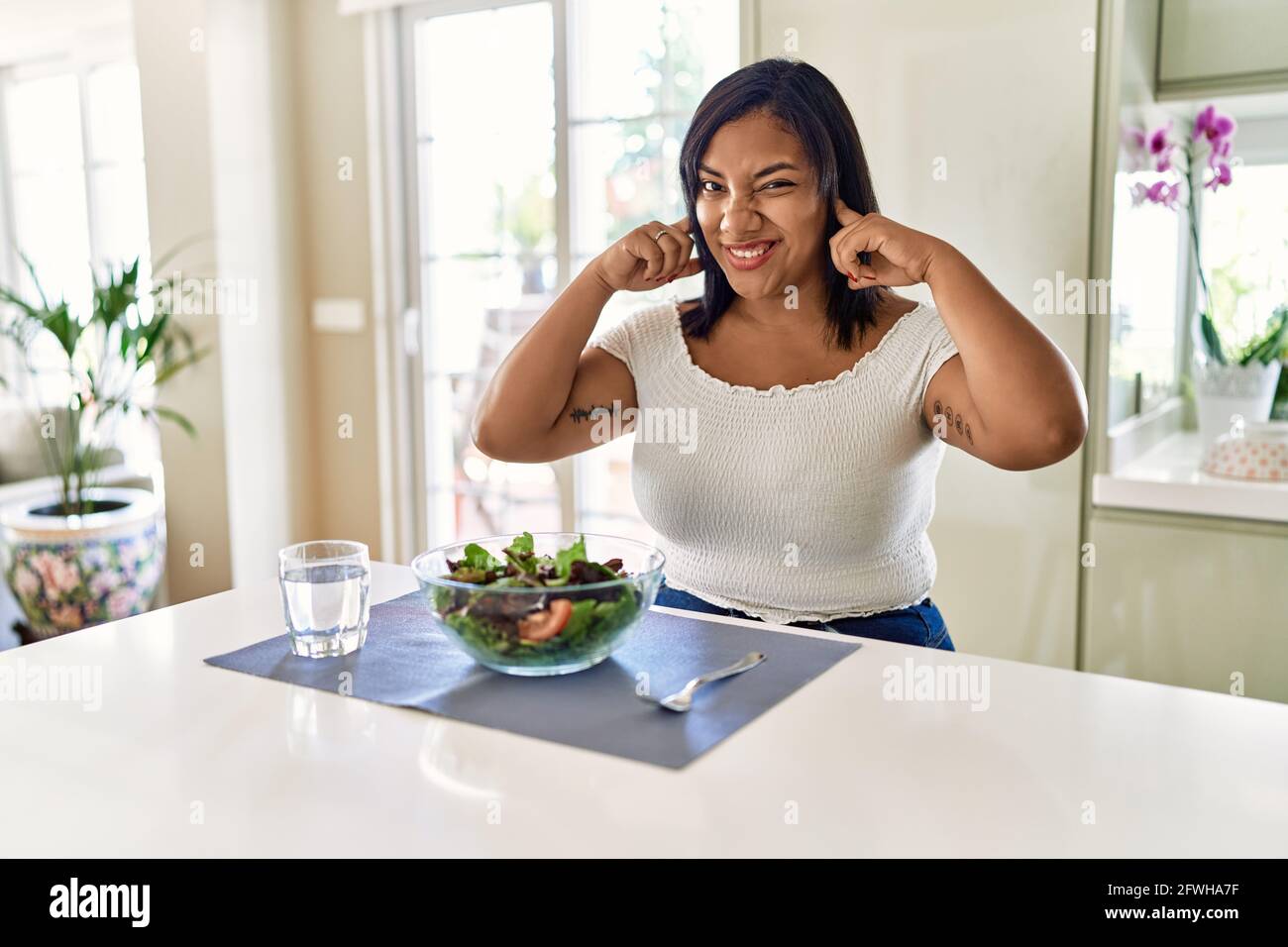 Young hispanic woman eating healthy salad at home covering ears with ...