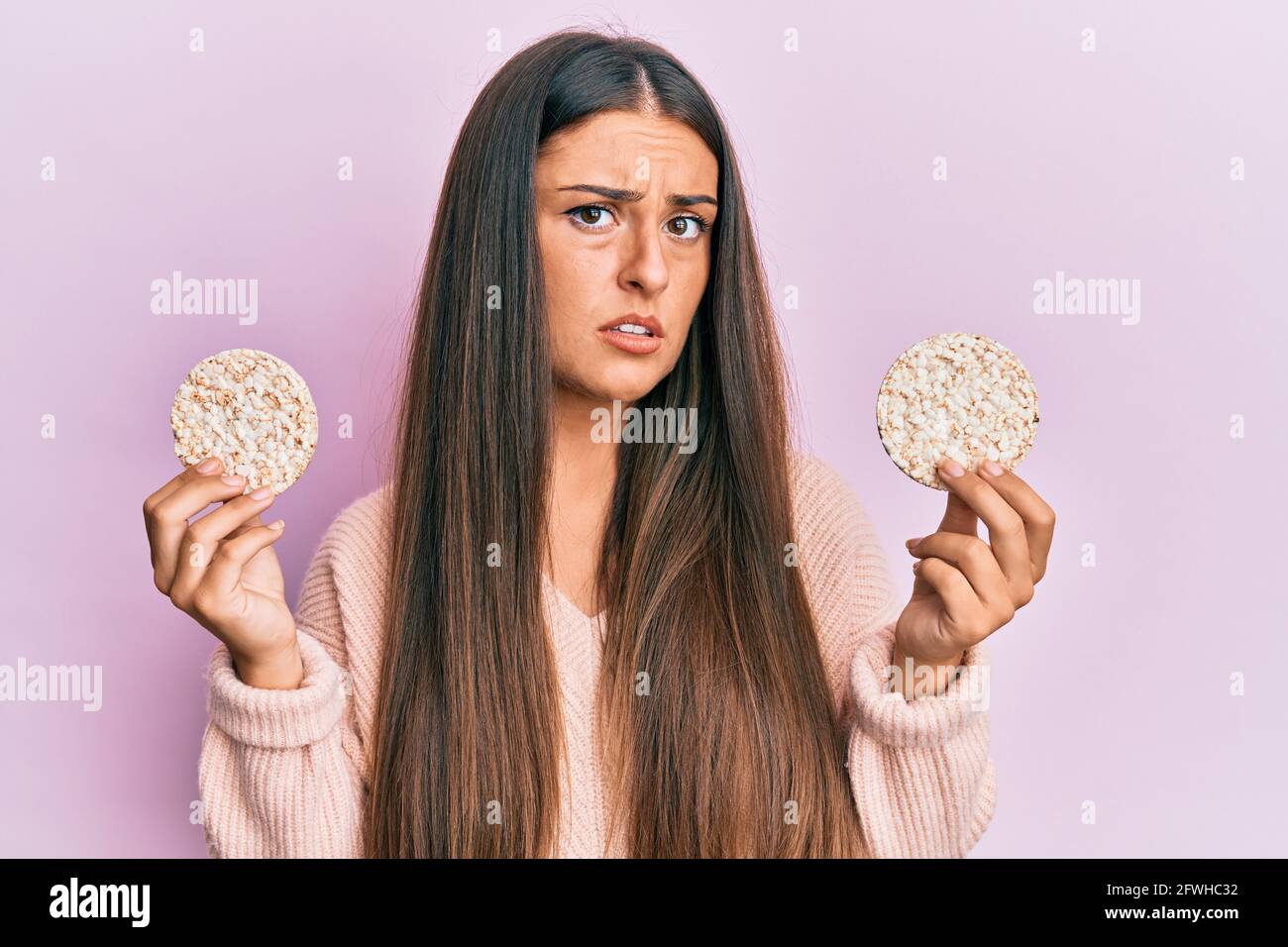Beautiful hispanic woman eating healthy rice crackers clueless and ...