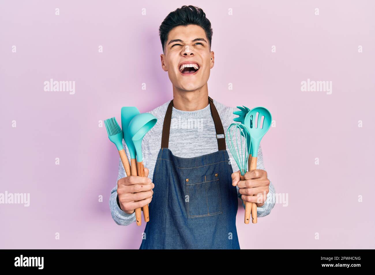 Young hispanic man wearing professional baker apron holding cooking tools angry and mad ...