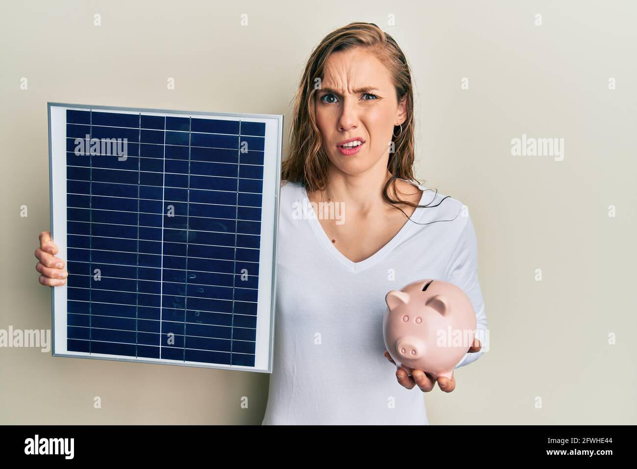 Young blonde woman holding photovoltaic solar panel and piggy bank ...