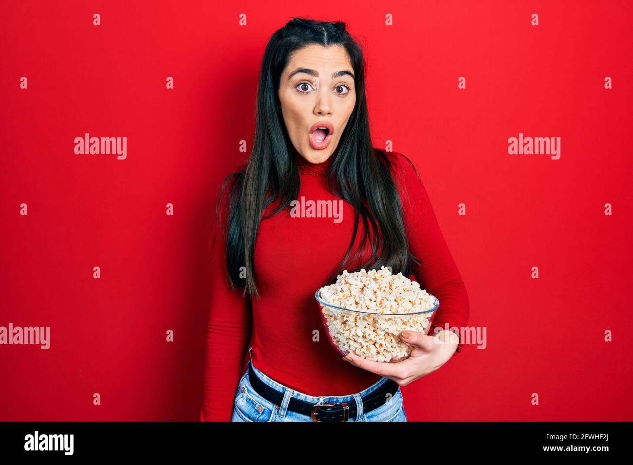 Young hispanic girl eating popcorn scared and amazed with open mouth ...