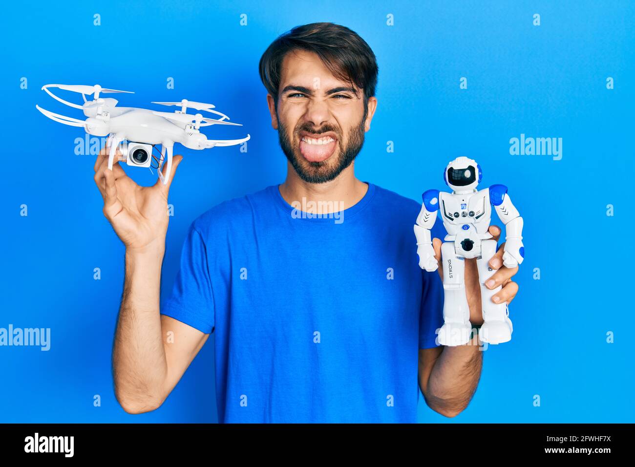 Young Hispanic Man Holding Drone And Robot Toy Sticking Tongue Out