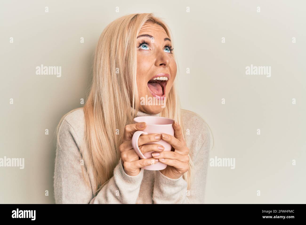 Young blonde woman drinking a cup of coffee angry and mad screaming ...