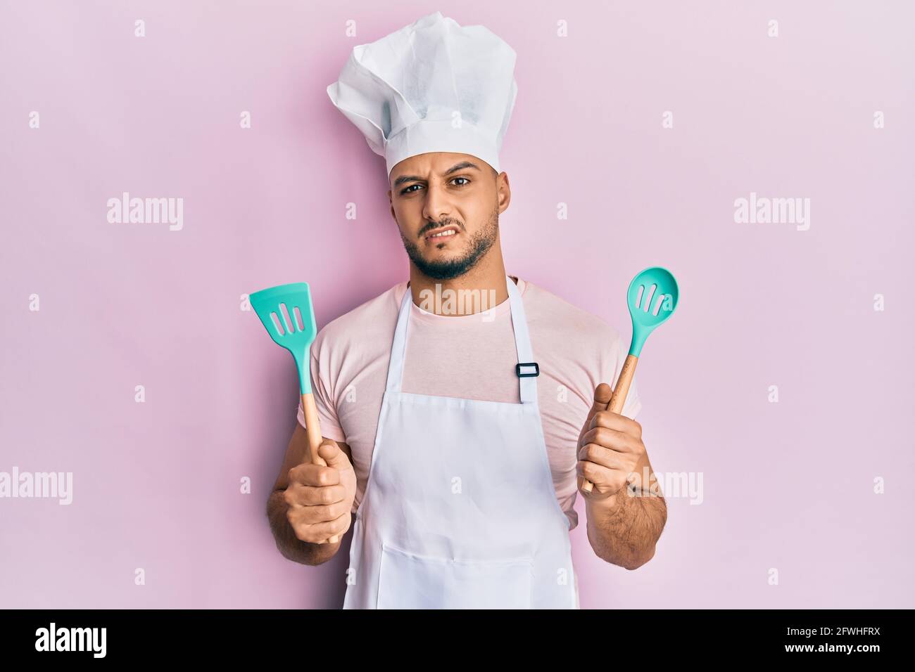 Young arab man wearing professional cook apron and hat holding spoon ...