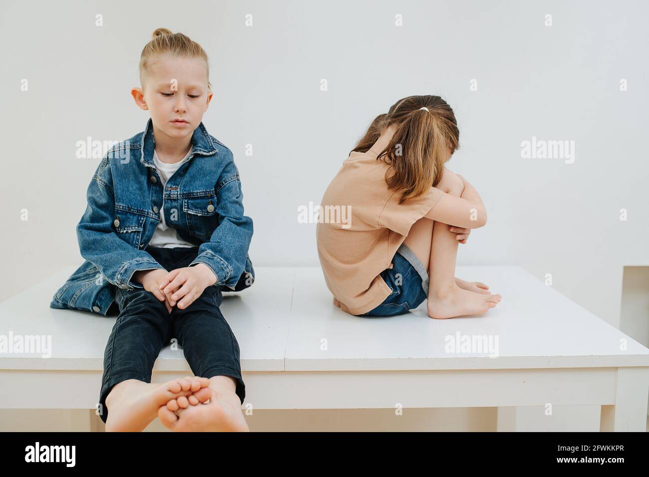 Sad little siblings sitting on a table. Boy has offended girl. He is ...