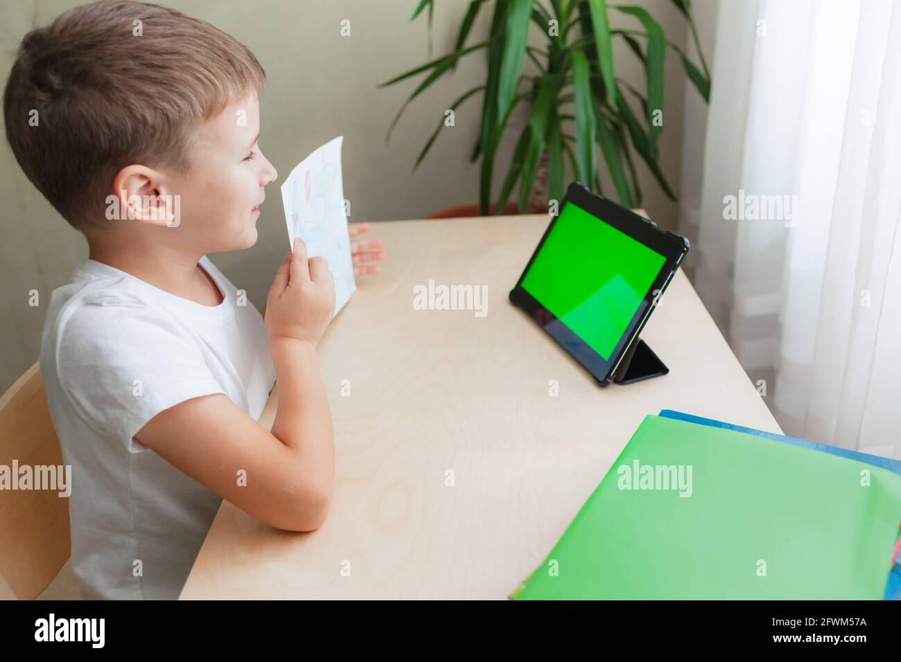 Smiling school boy siting at desk and looks at tablet with green screen ...
