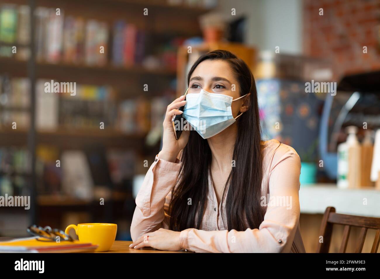 Arab lady wearing face mask and talking on smartphone, sitting at table ...