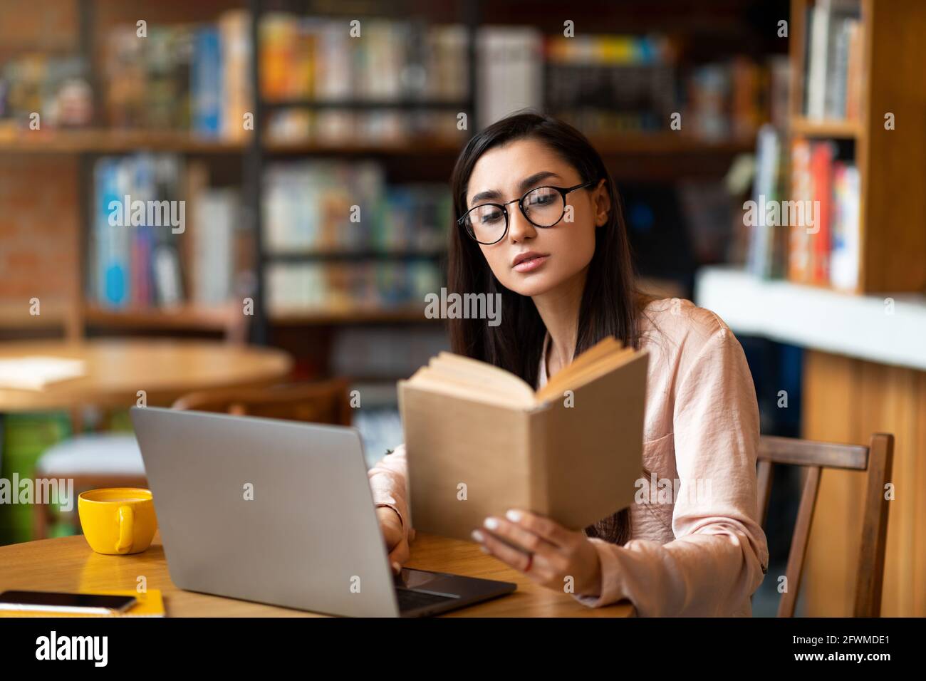 Thoughtful latin woman reading book in front of laptop at cafeteria ...