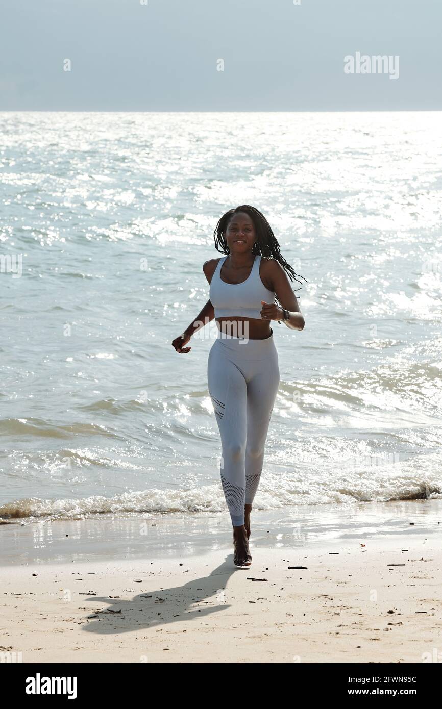 Happy fit beautiful sportswoman in activewear jogging on beach on sunny ...