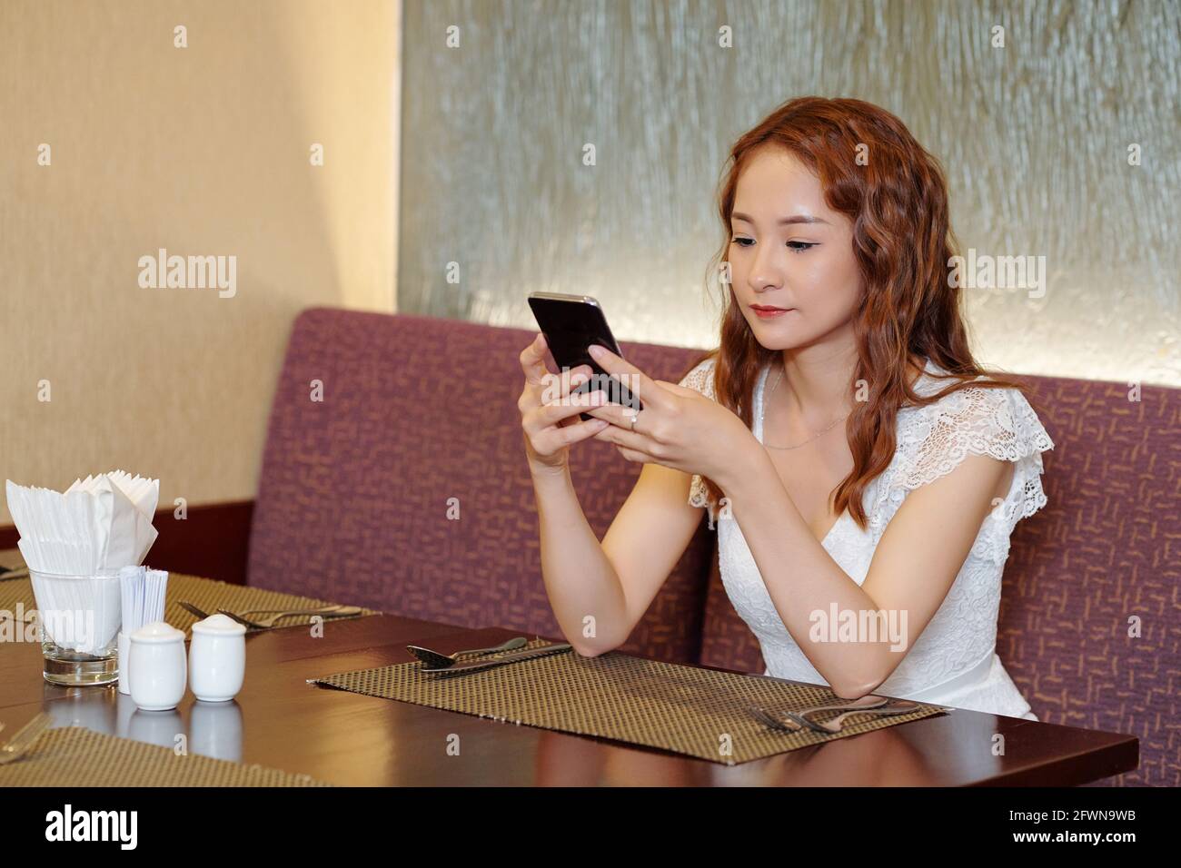 Pretty young woman sitting at restaurant table, texting friends or ...