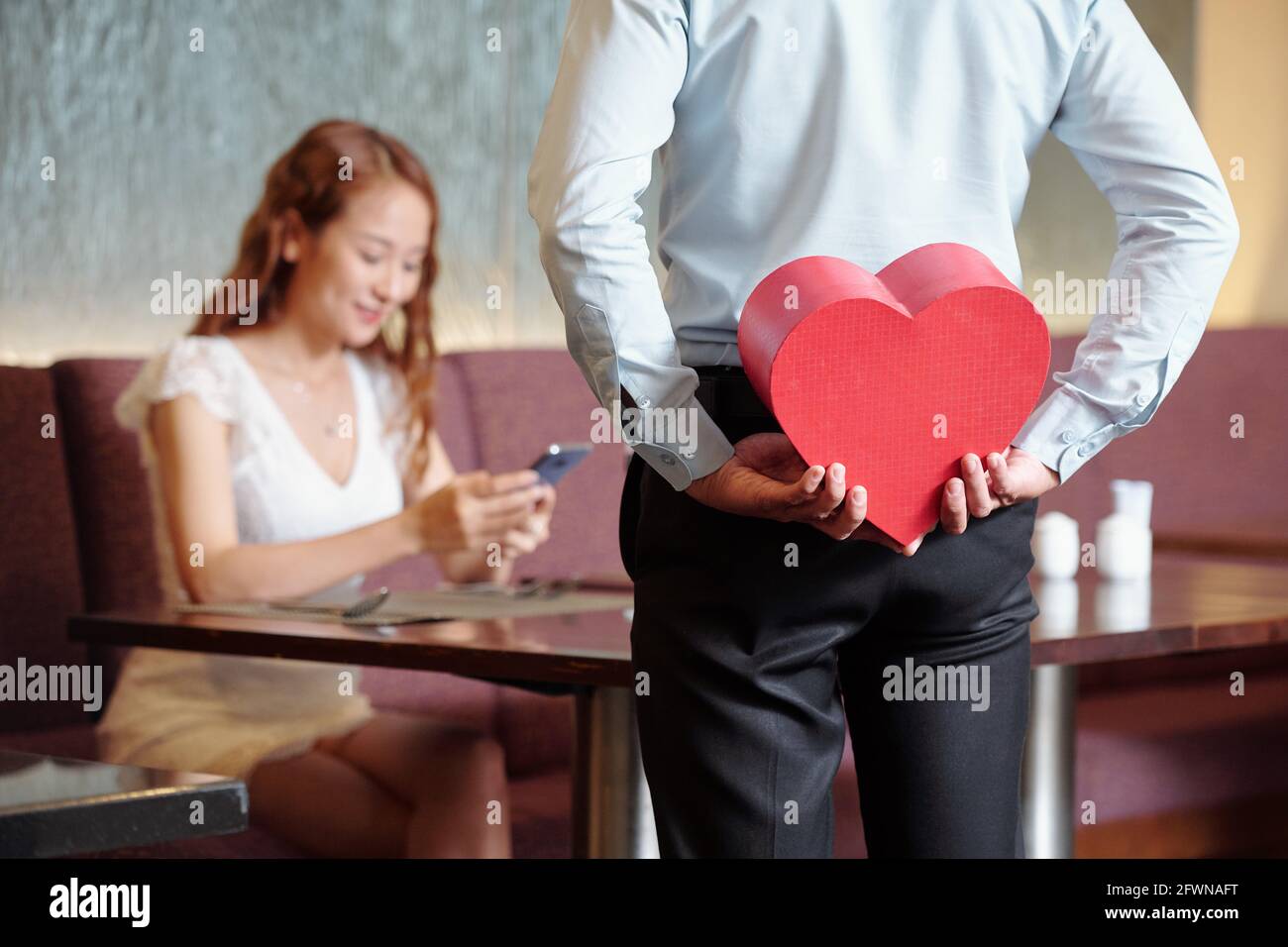 Man hiding present in heart shape box for girlfriend who is sitting at ...