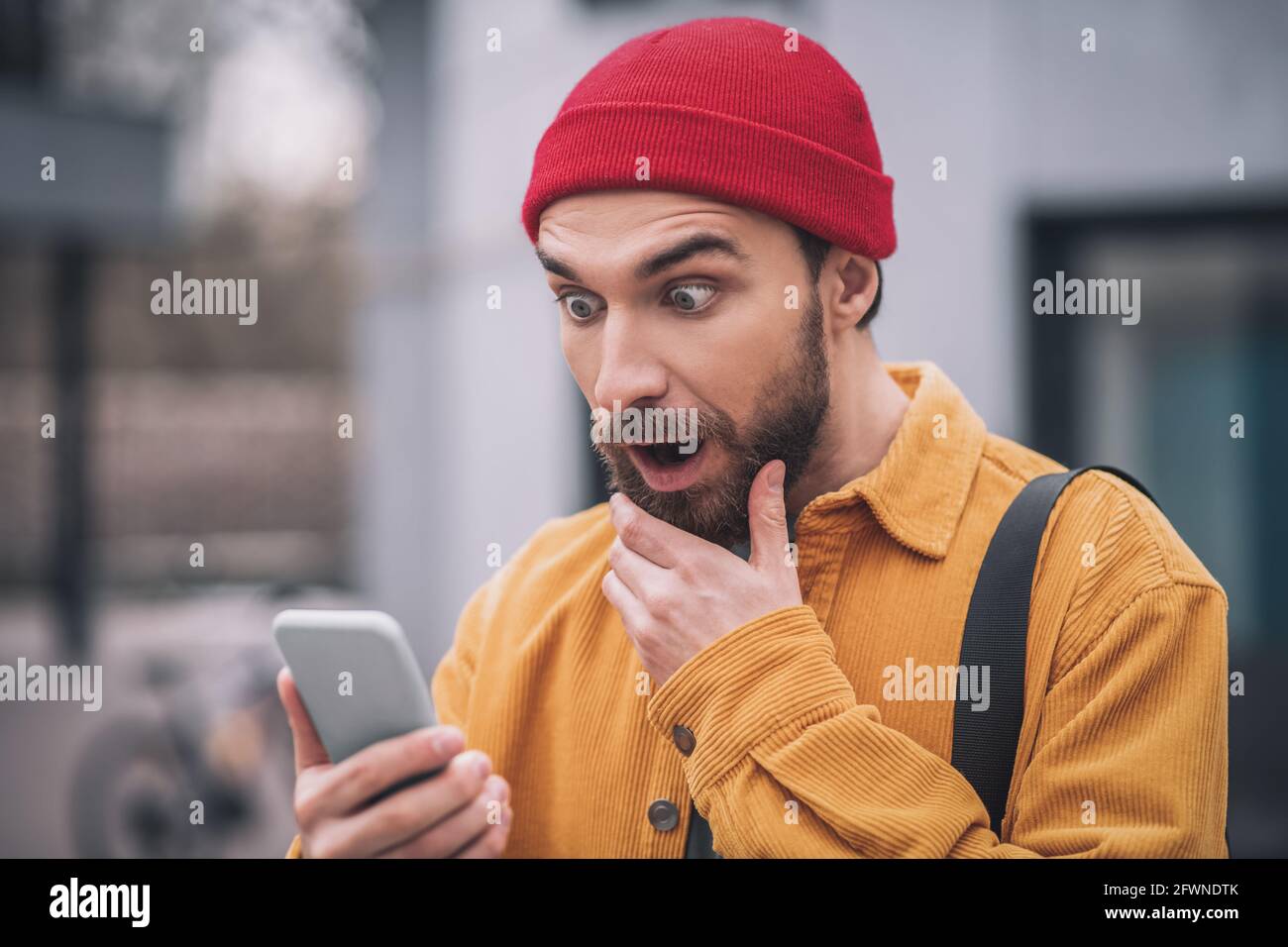 Young bearded man in a red hat looking at his phone and feeling happy ...