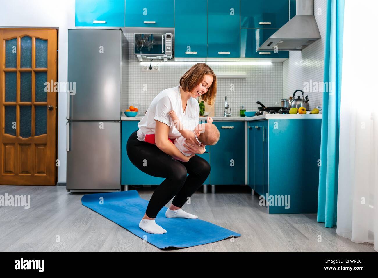 Young Caucasian mother squats in the kitchen, holding the baby in her ...