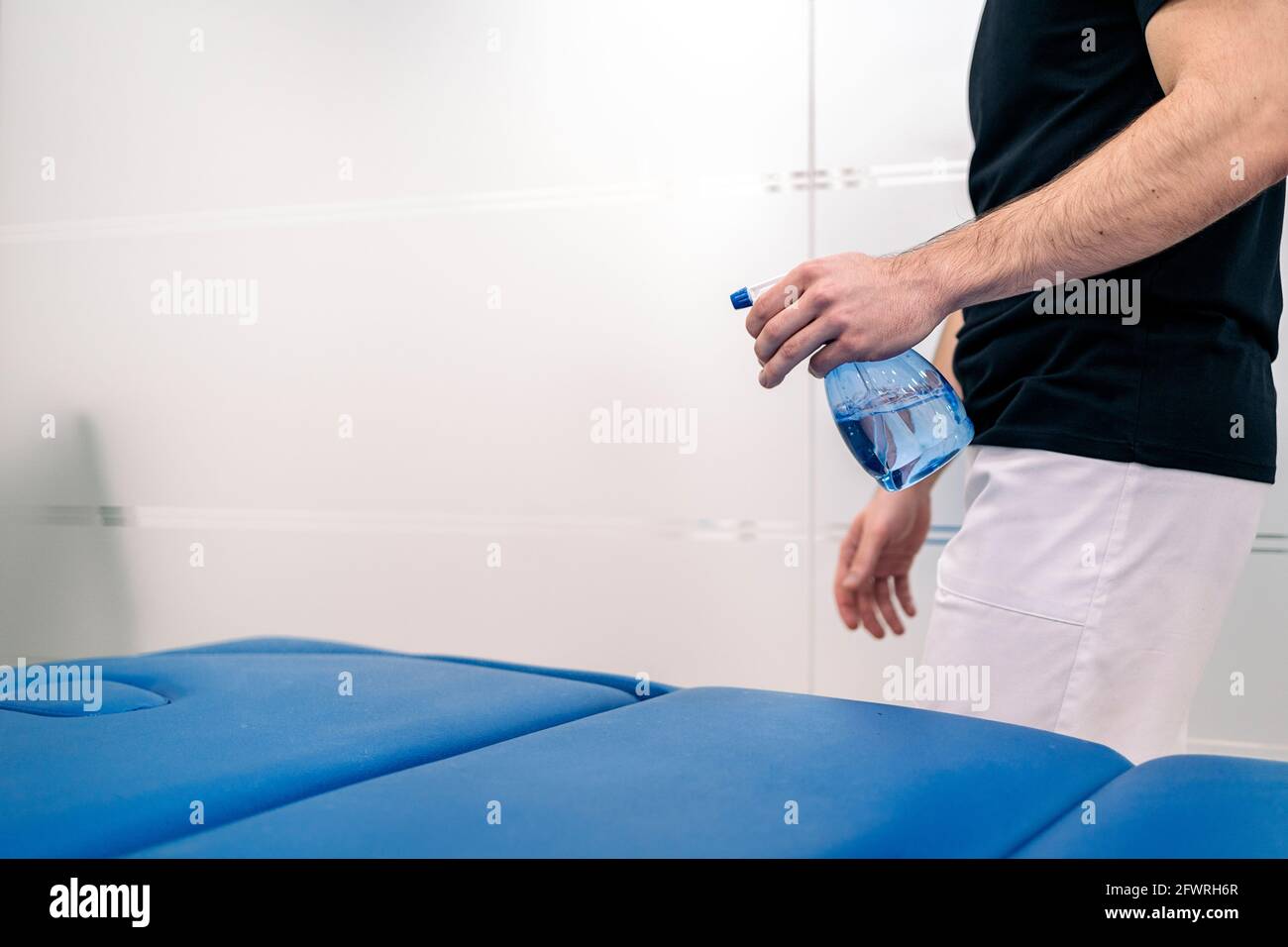 Male physiotherapist using spray with sanitizer to clean blue stretcher ...