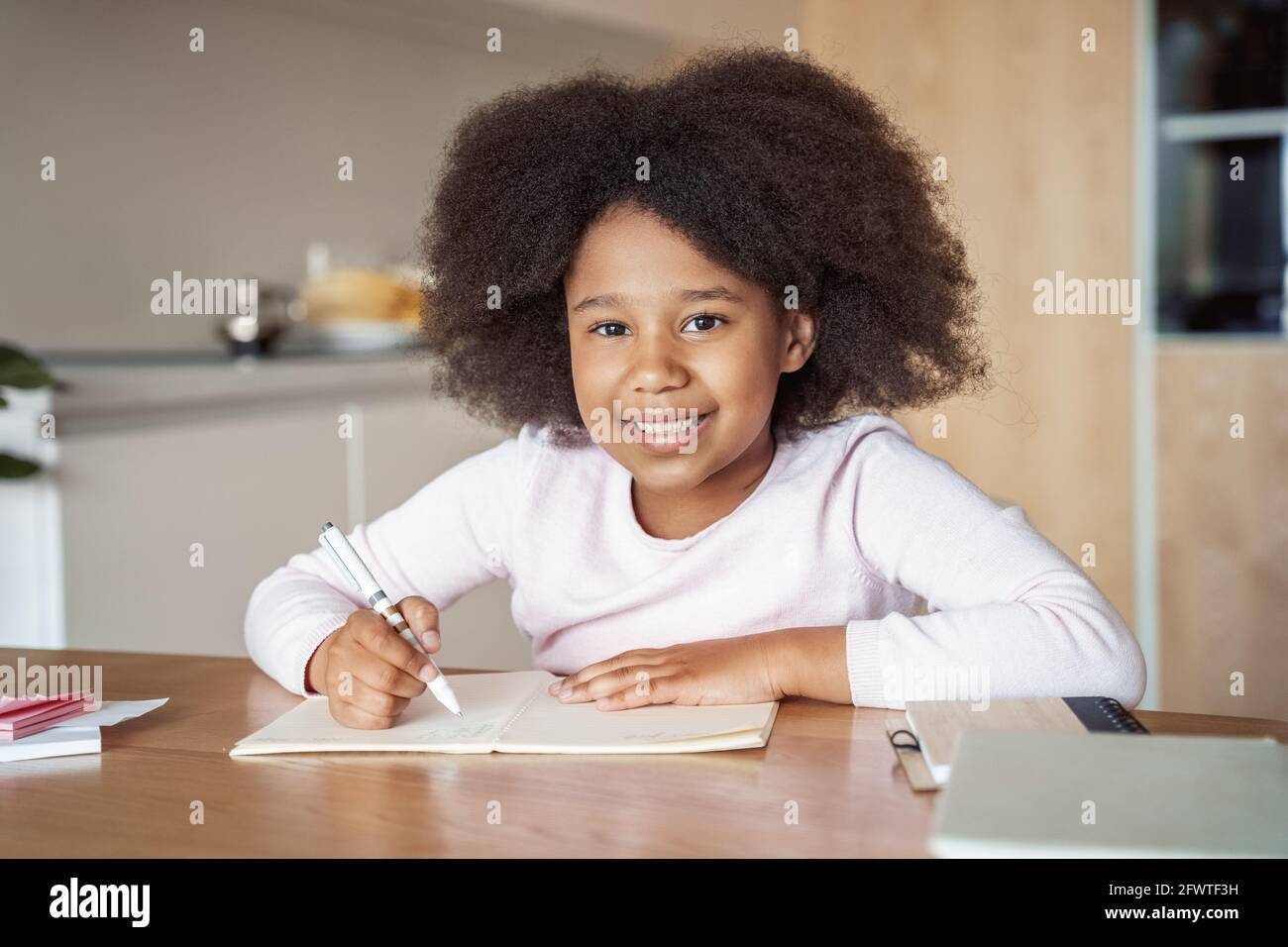 Happy little mixed-race female child writing homework with pen smiling ...