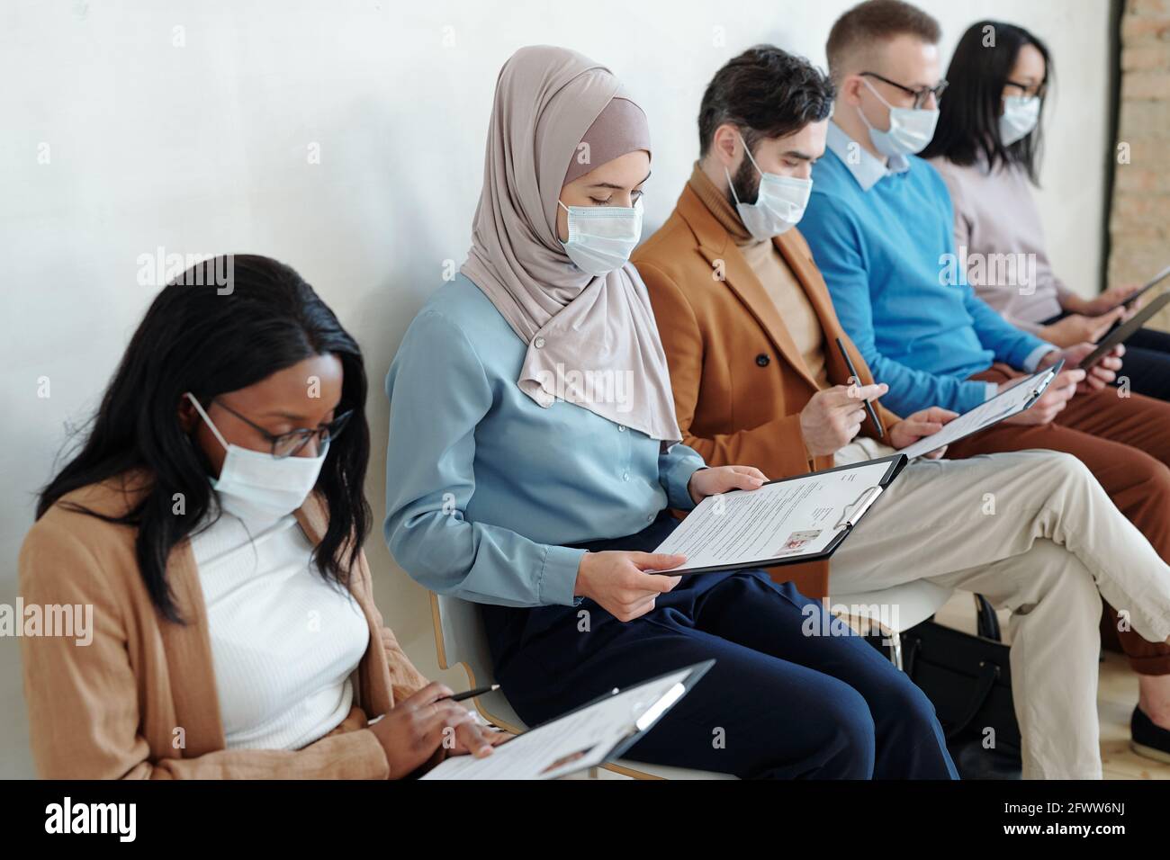 Group of young multi-ethnic job candidates in masks sitting in row and ...