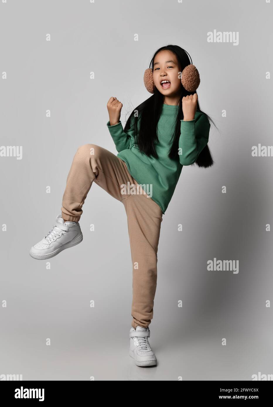 Asian kid girl in green earmuffs, sportswear and white sneakers stands ...