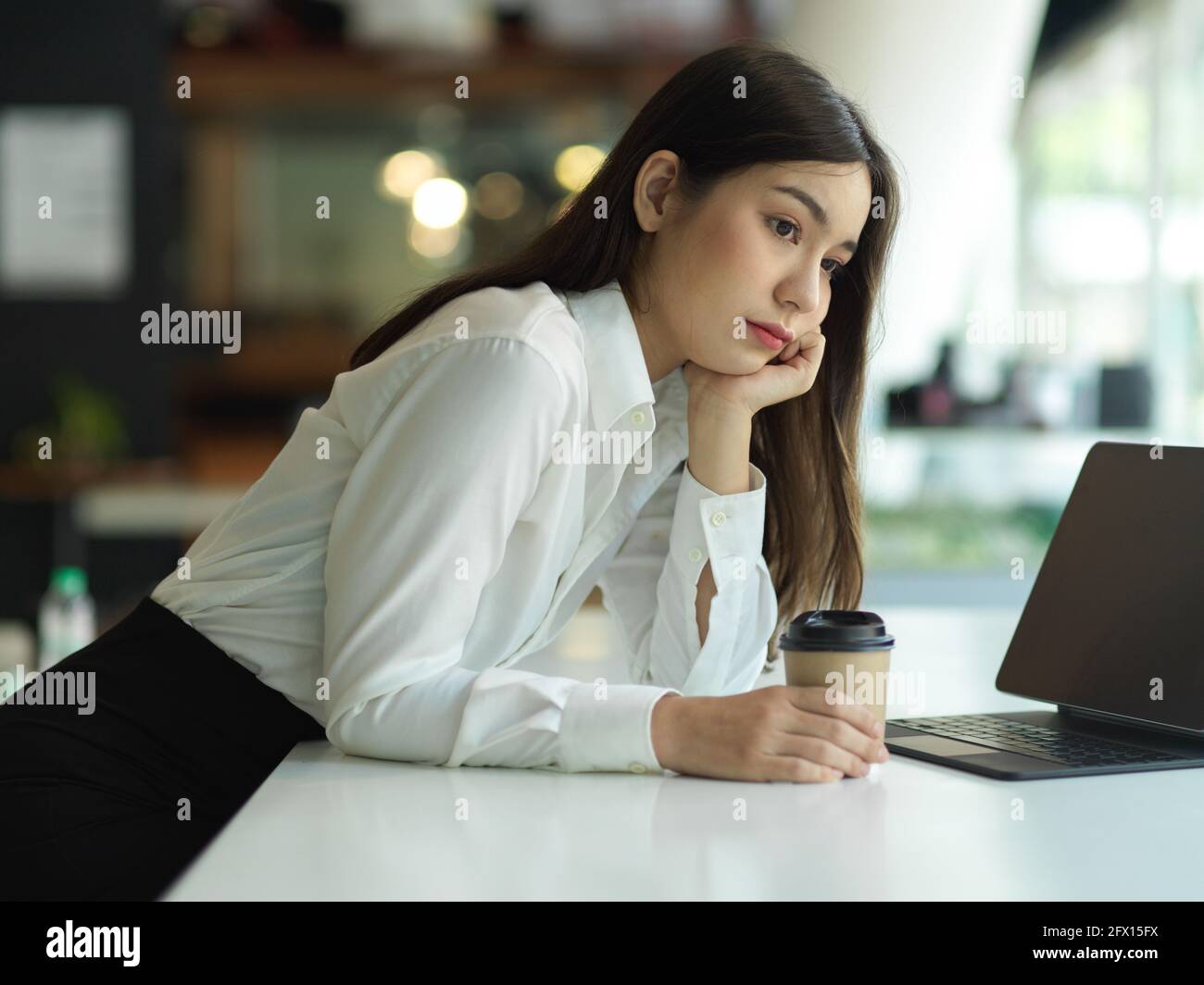 Side view of female worker sitting hand to cheek and holding paper cup ...