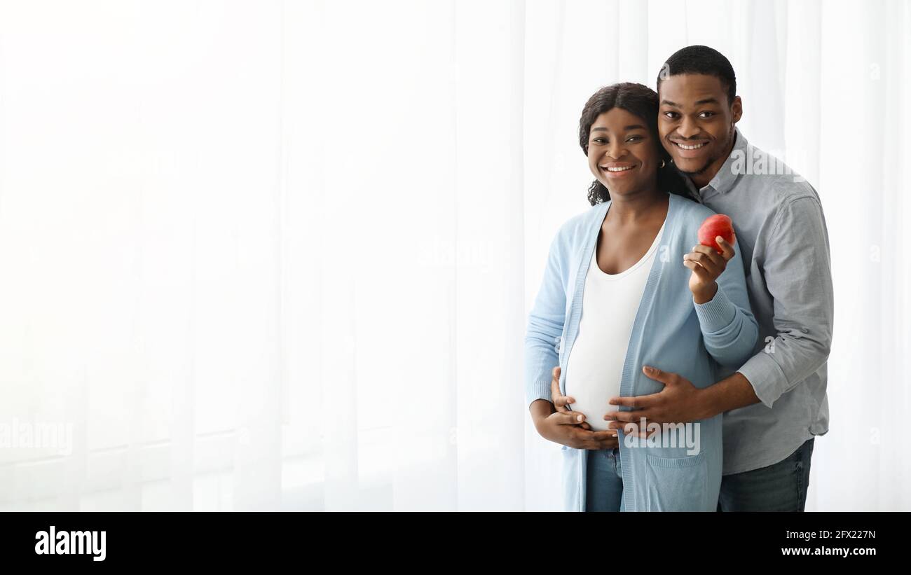 Cheerful pregnant black family cuddling, showing red apple Stock Photo ...