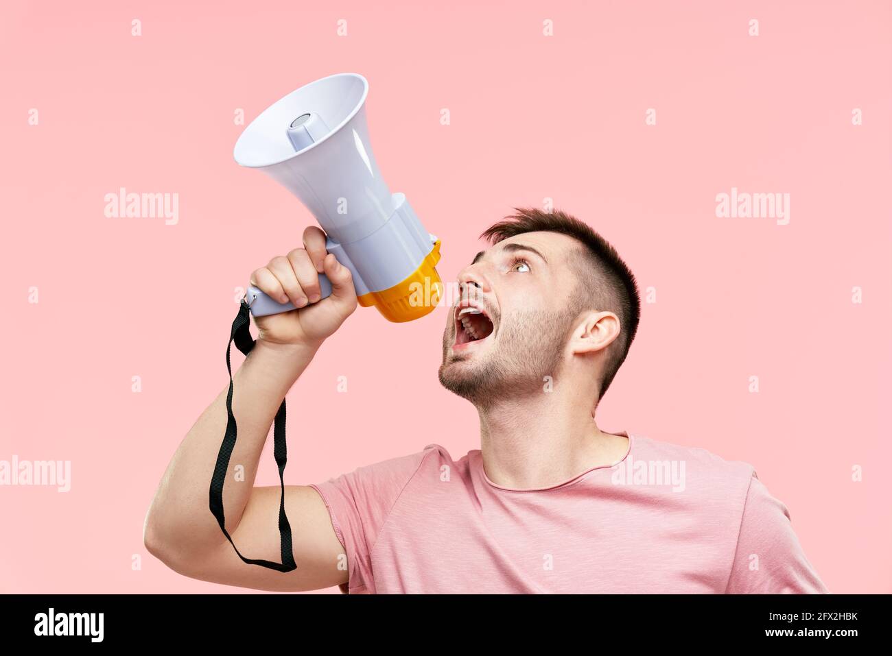 Funny young man shouting loud holding a megaphone over pink background ...