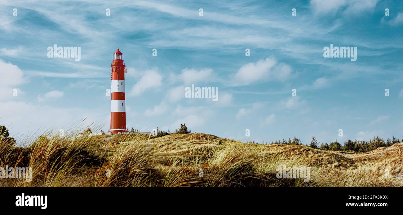 Tall red and white striped lighthouse on sand dunes in evening sunlight ...