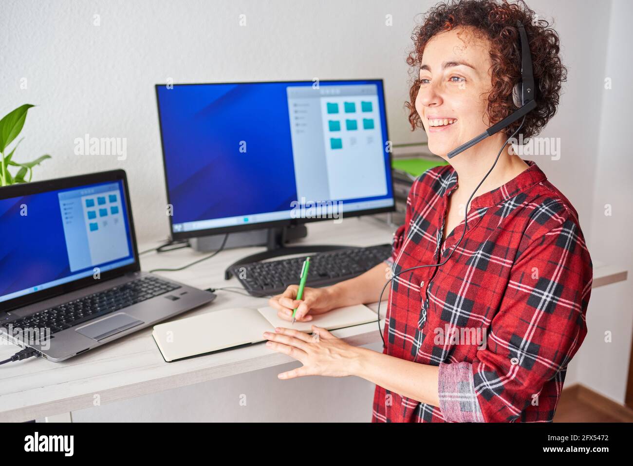 Woman working remotely standing up, wearing headset and taking notes on ...