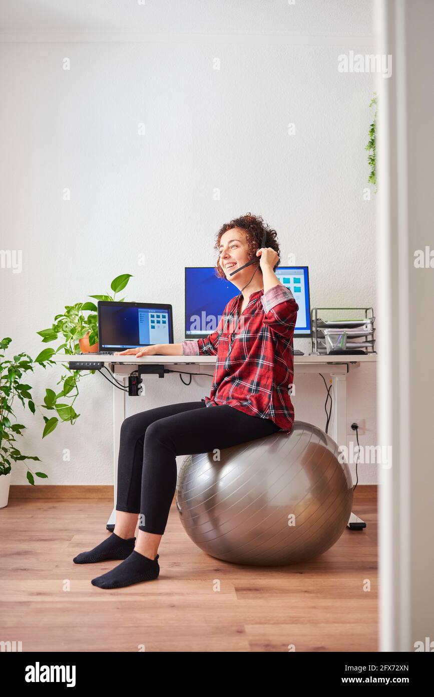 Woman teleworking sitting on a fitball in front of her desk talking on ...