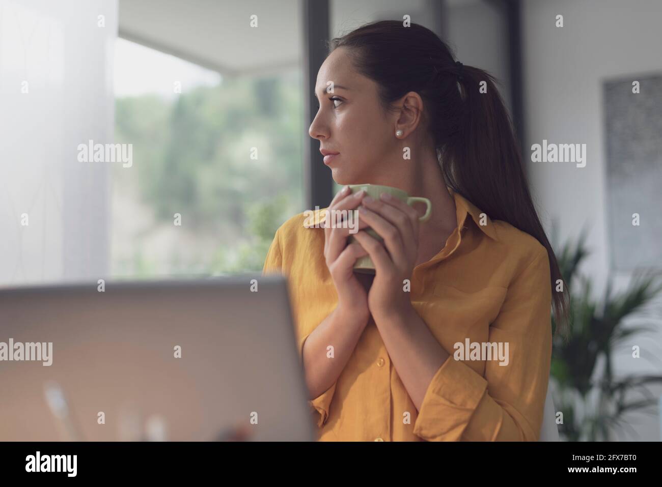 Young woman sitting at office desk and drinking coffee, she is looking ...