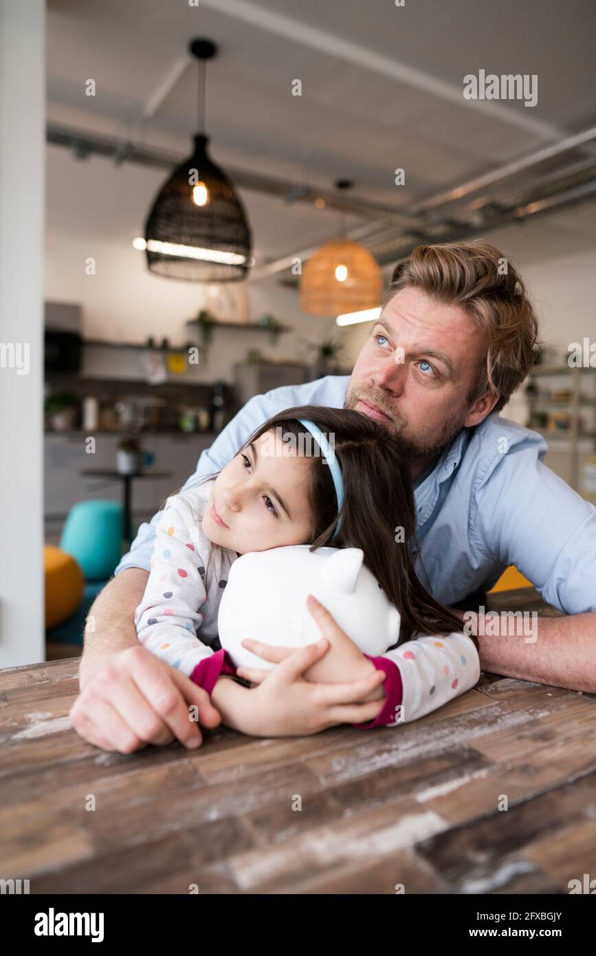Thoughtful father and daughter with piggy bank looking away at home ...