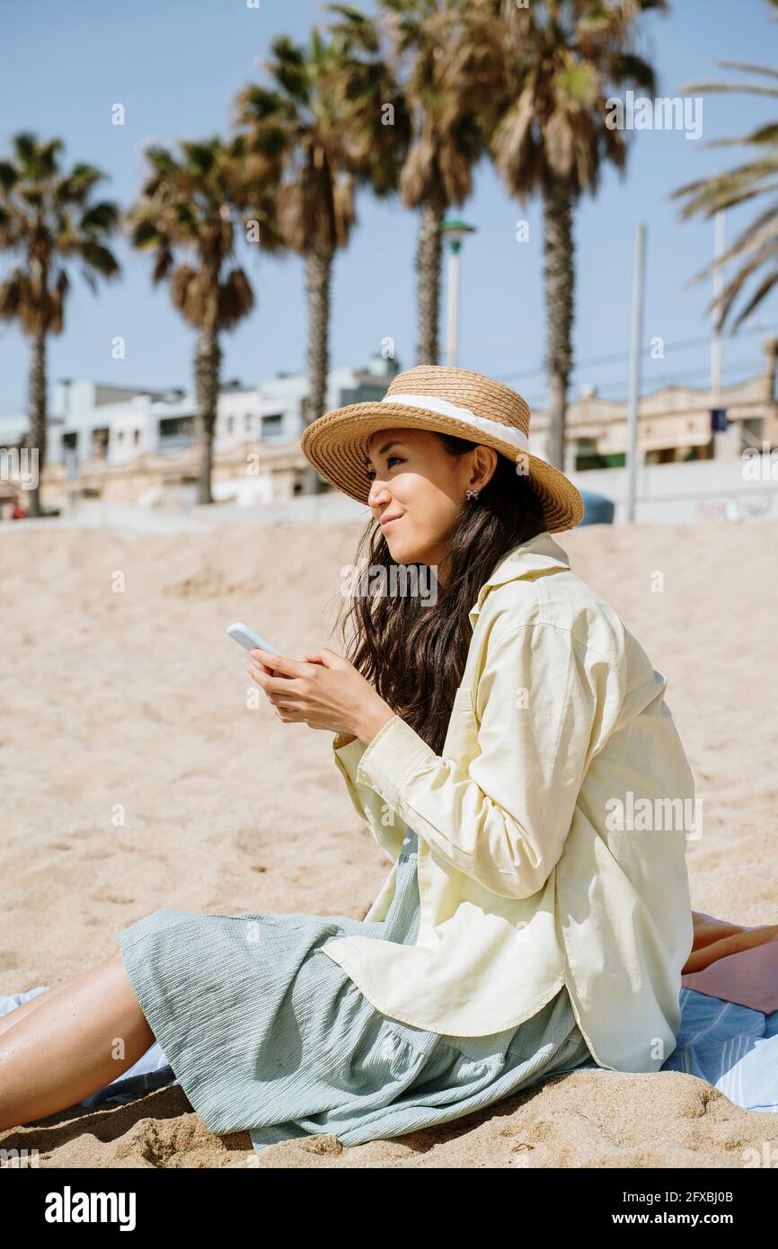 Smiling woman looking away while holding smart phone on beach during ...