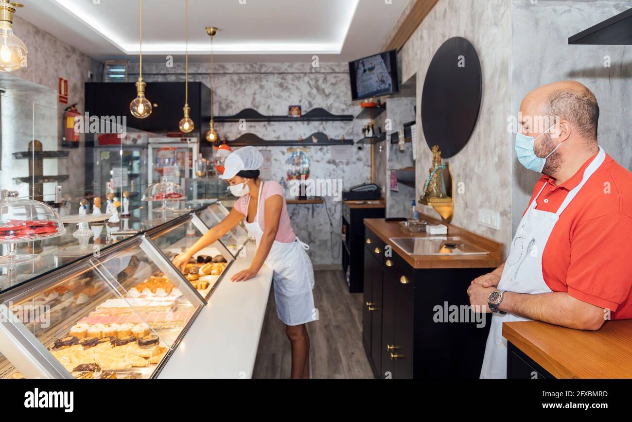 Male owner wearing face mask looking at woman arranging pastry in ...