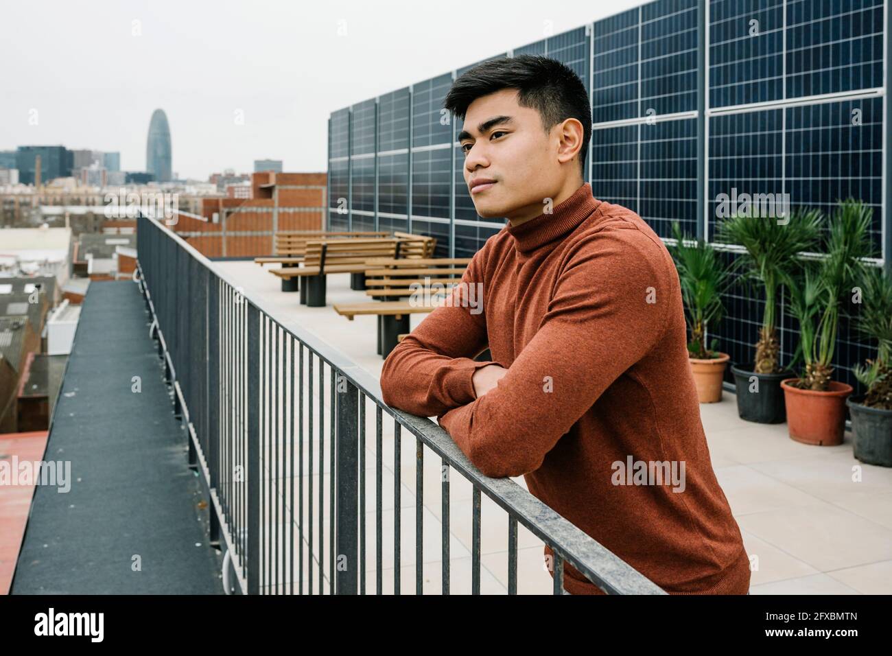 Thoughtful businessman with arms crossed leaning on railing at rooftop ...