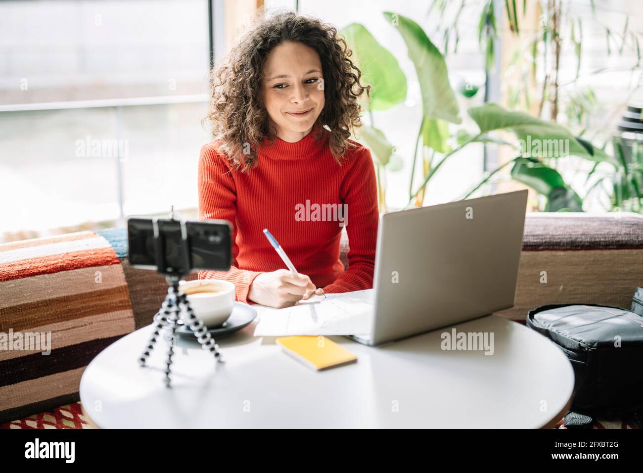 Smiling young female influencer writing in book while vlogging through ...
