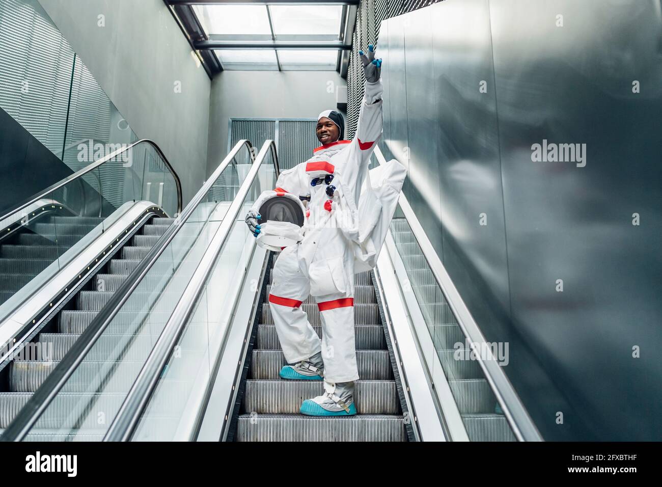 Cheerful astronaut gesturing peace sign while climbing on escalator ...