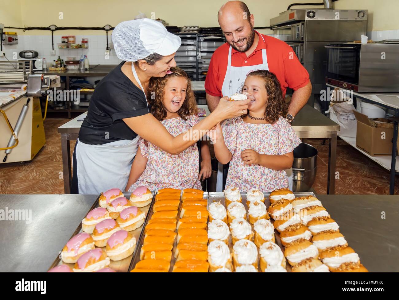 Smiling mother feeding cake to girls while standing with man in bakery ...