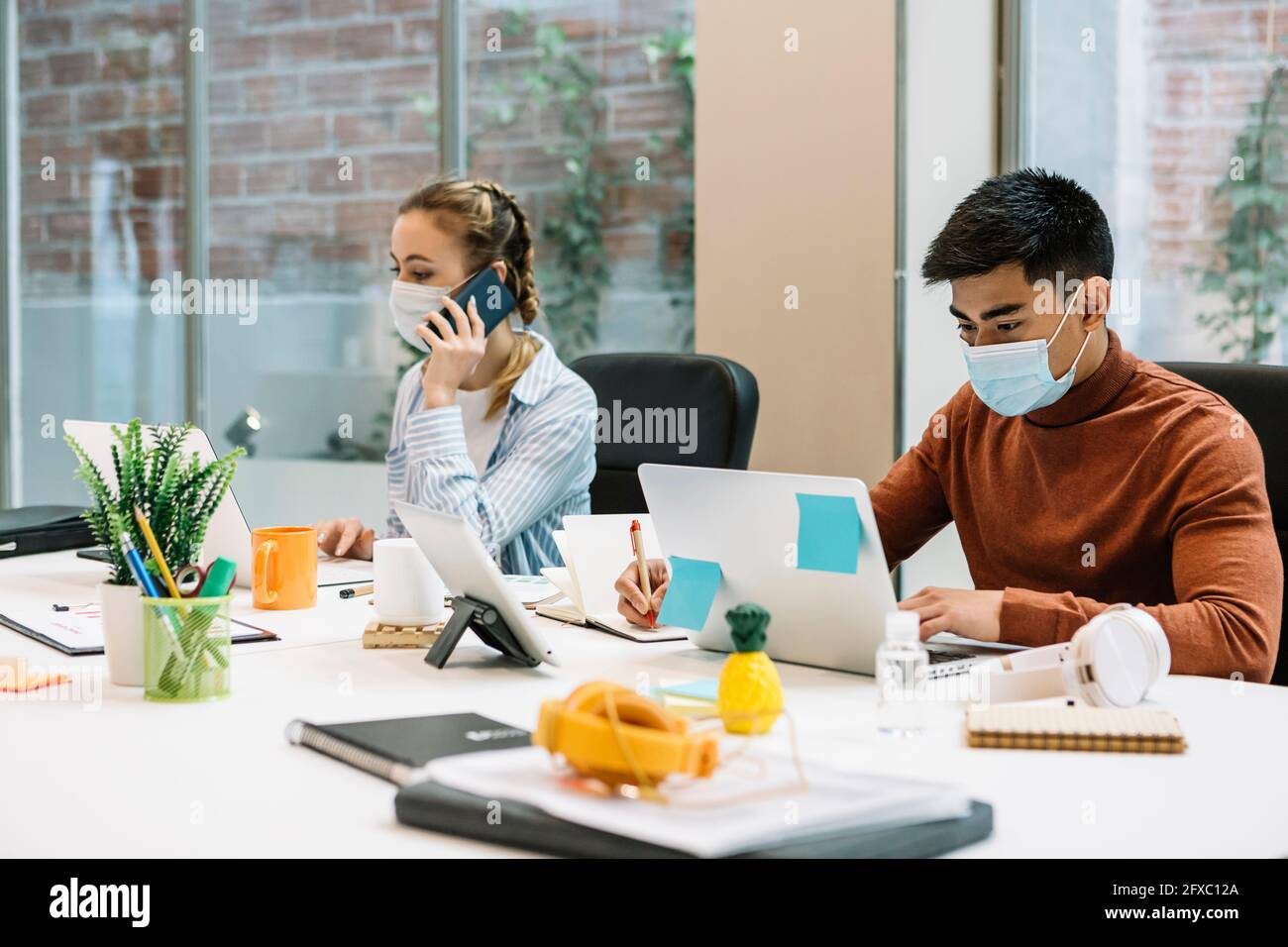 Male and female professionals using laptop at office during COVID-19 ...