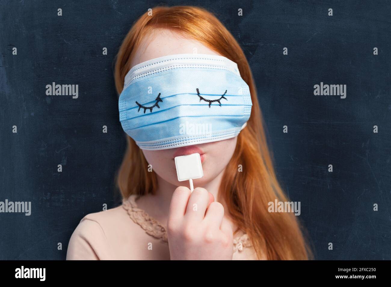 Girl covering eyes with protective face mask while eating lollipop ...