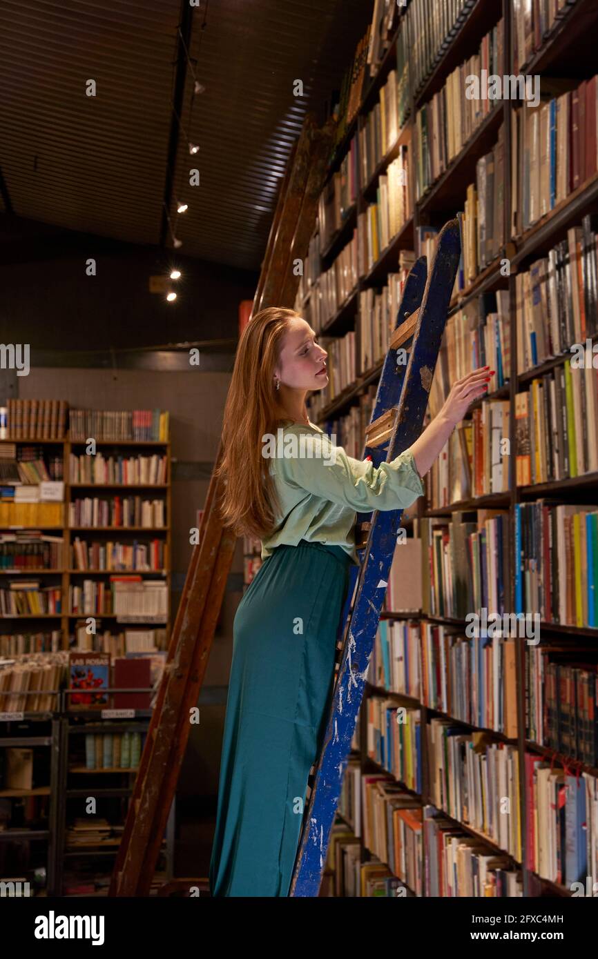 Woman with long hair standing on ladder while searching book in library ...