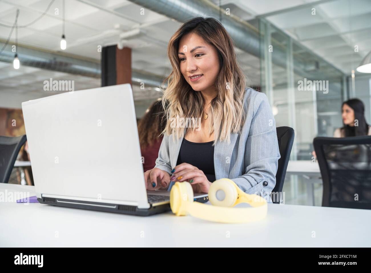 Smiling female entrepreneur using laptop at desk in coworking office ...
