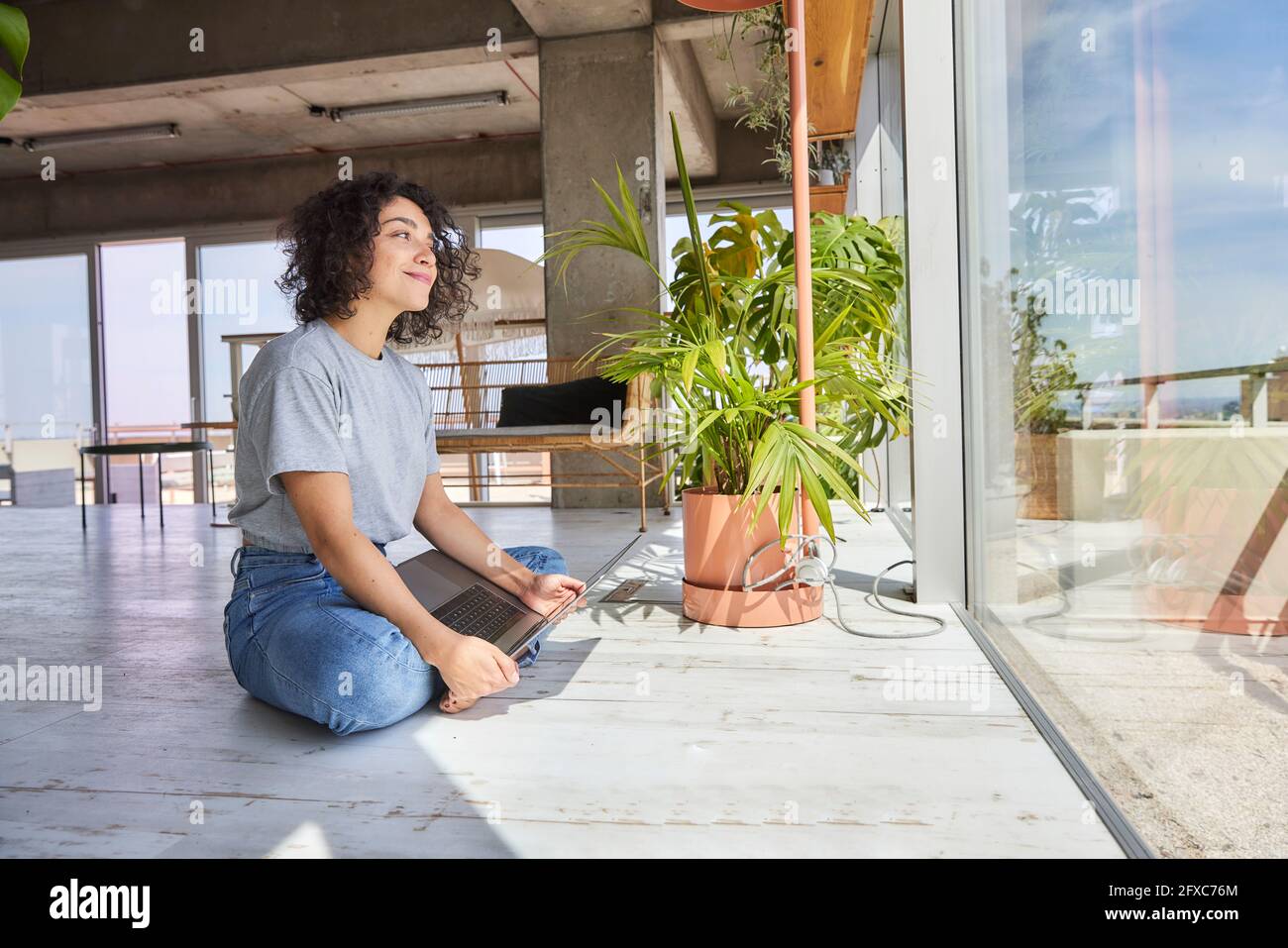 Woman looking through window while sitting on floor with laptop at home ...