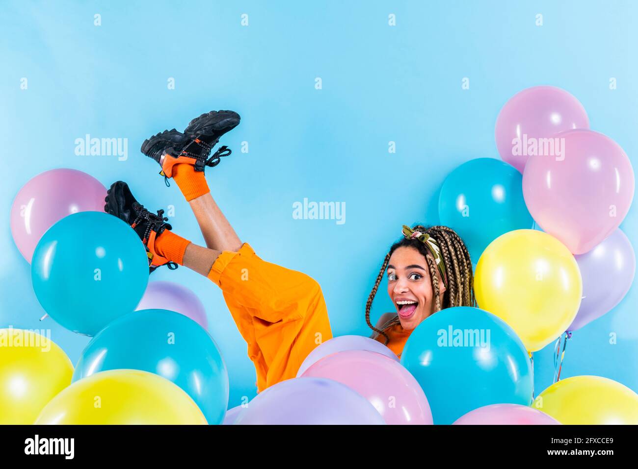 Playful young woman with feet up amidst balloons over blue background ...