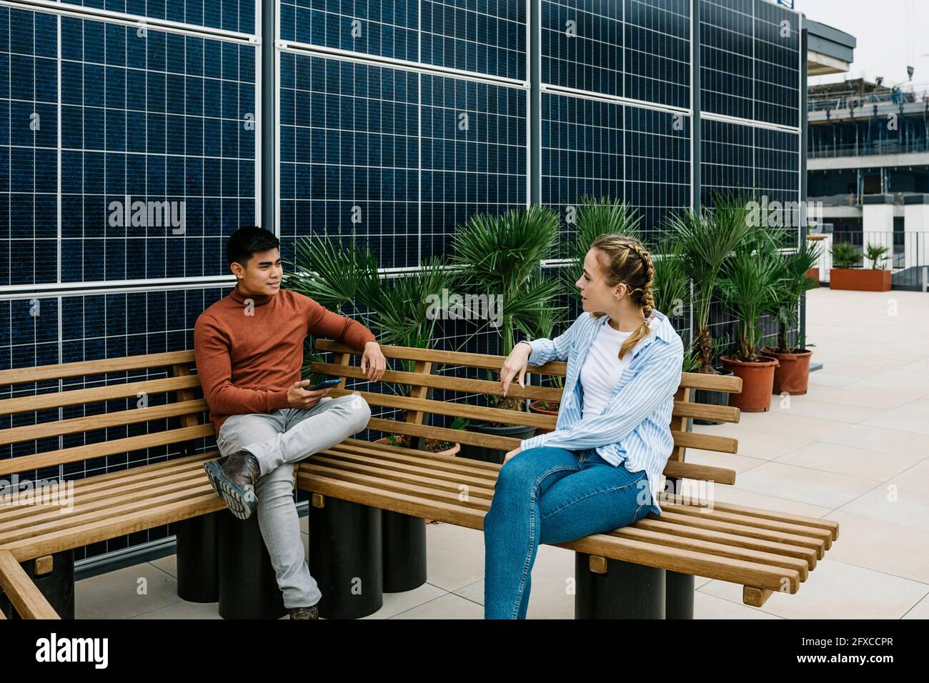 Male and female professionals talking while sitting on bench at rooftop ...