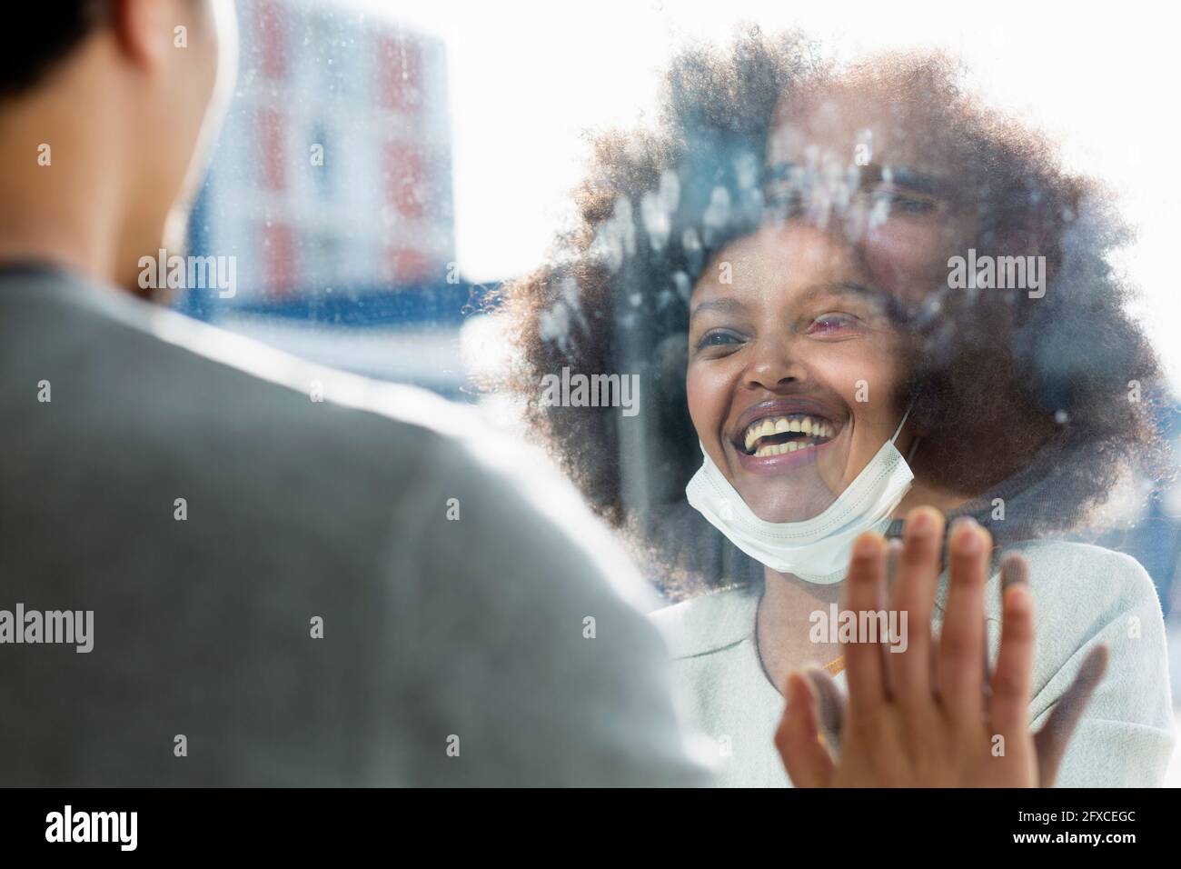 Smiling afro woman with face mask meeting male friend through glass ...