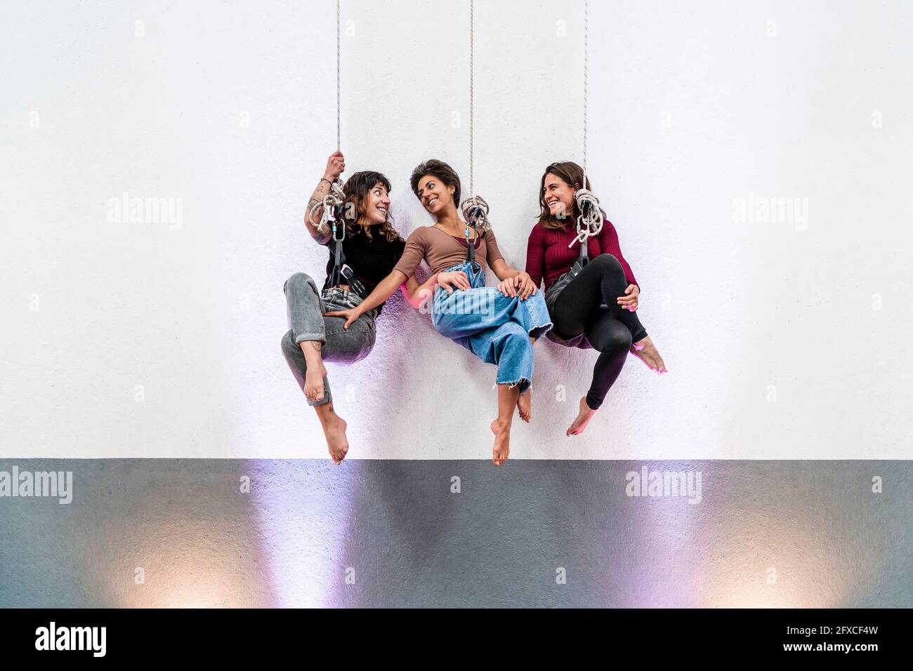 Smiling female acrobats hanging side by side on rope in front of wall ...