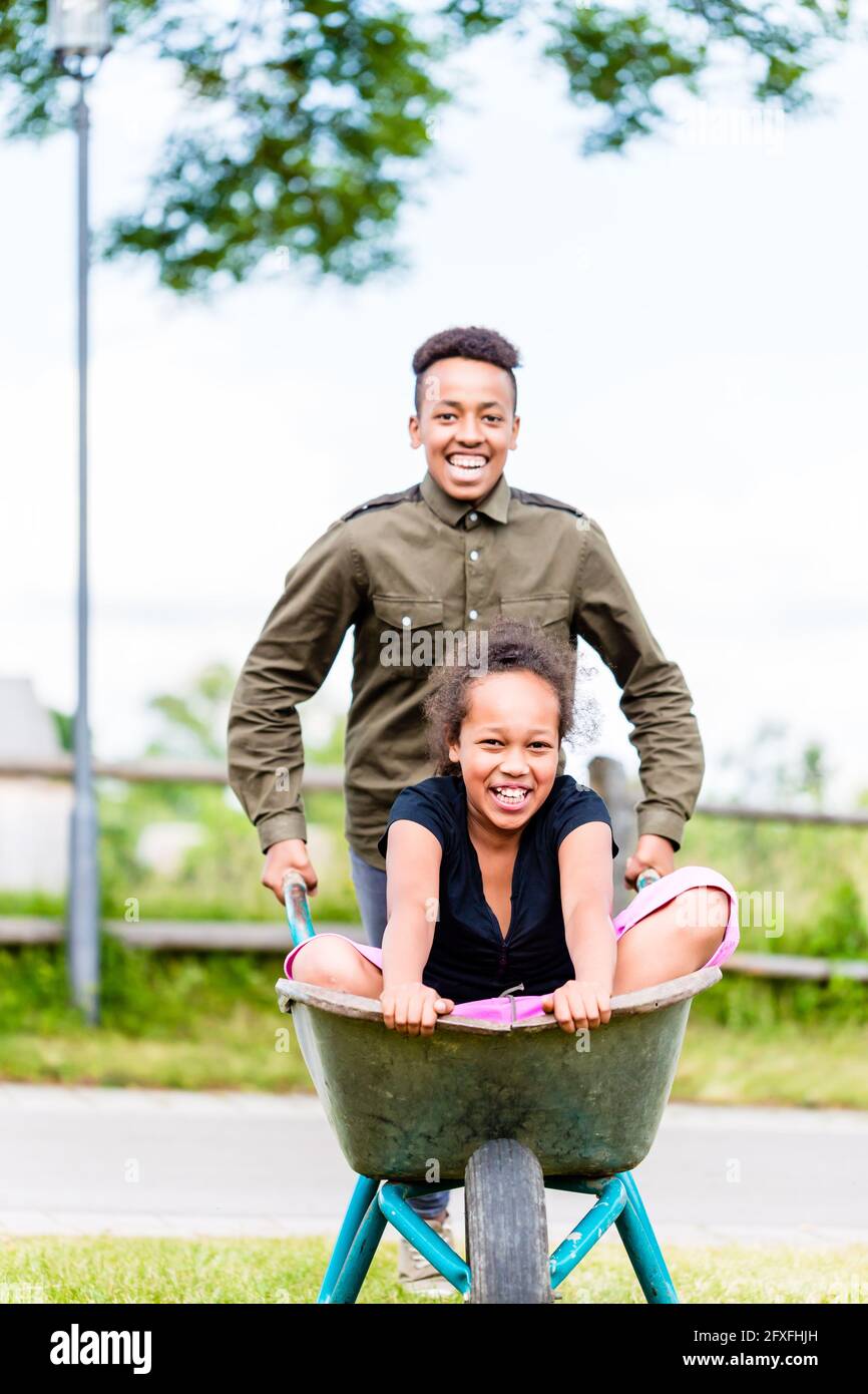 Siblings playing together in garden pushing each other in wheelbarrow ...