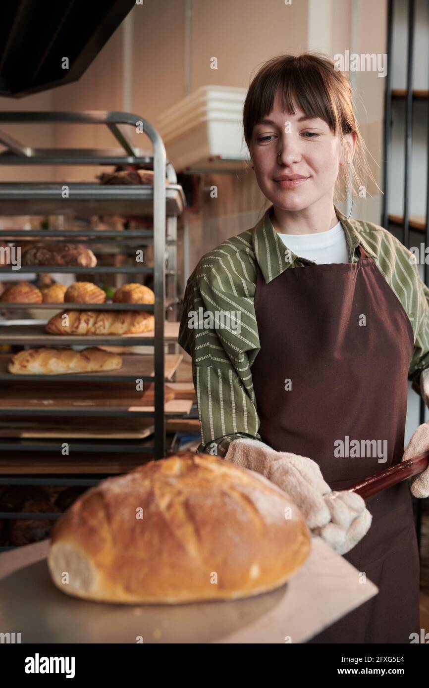Baker in uniform taking the bread out of the oven while working in ...