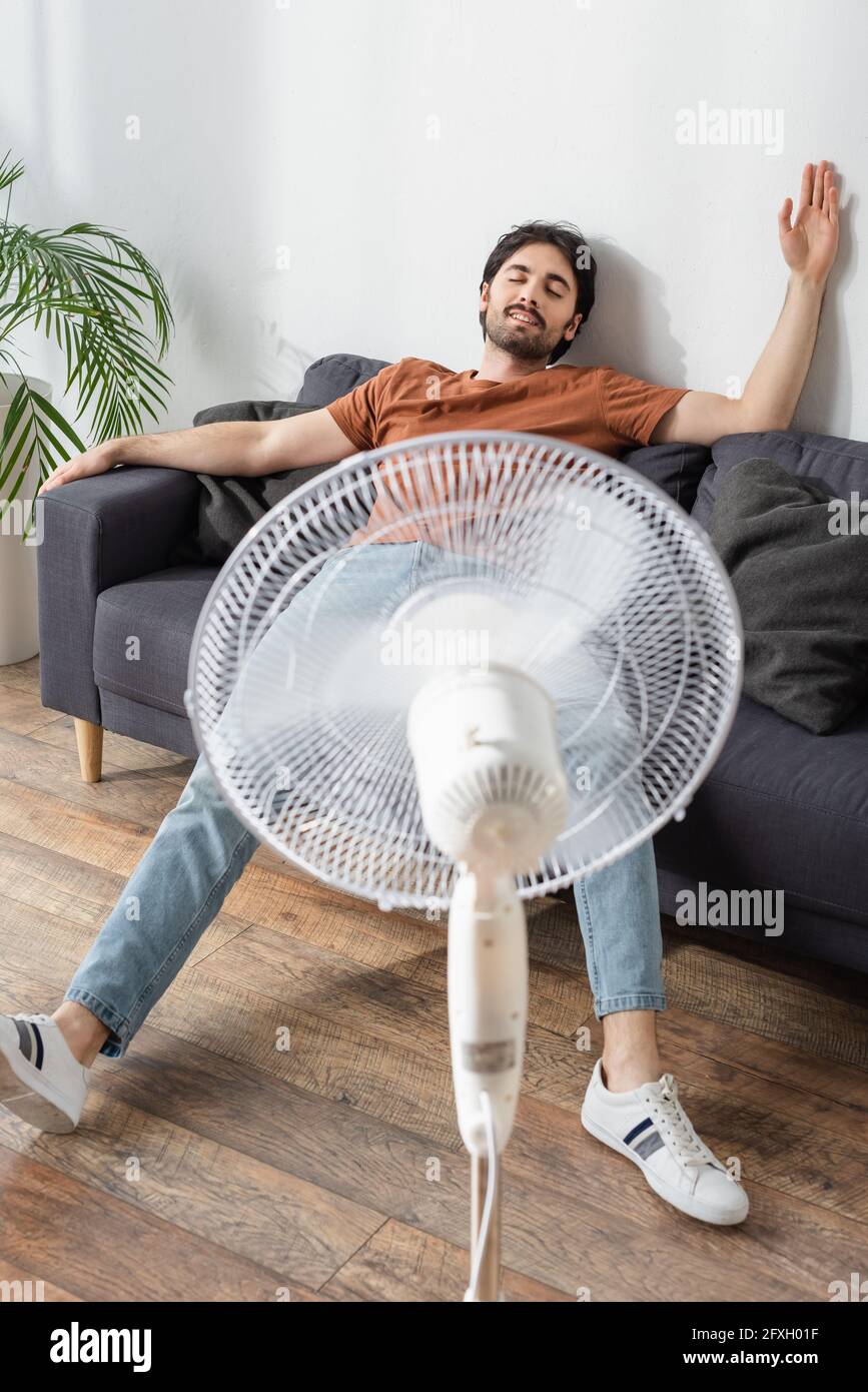 pleased man sitting on couch near blurred electric fan Stock Photo - Alamy