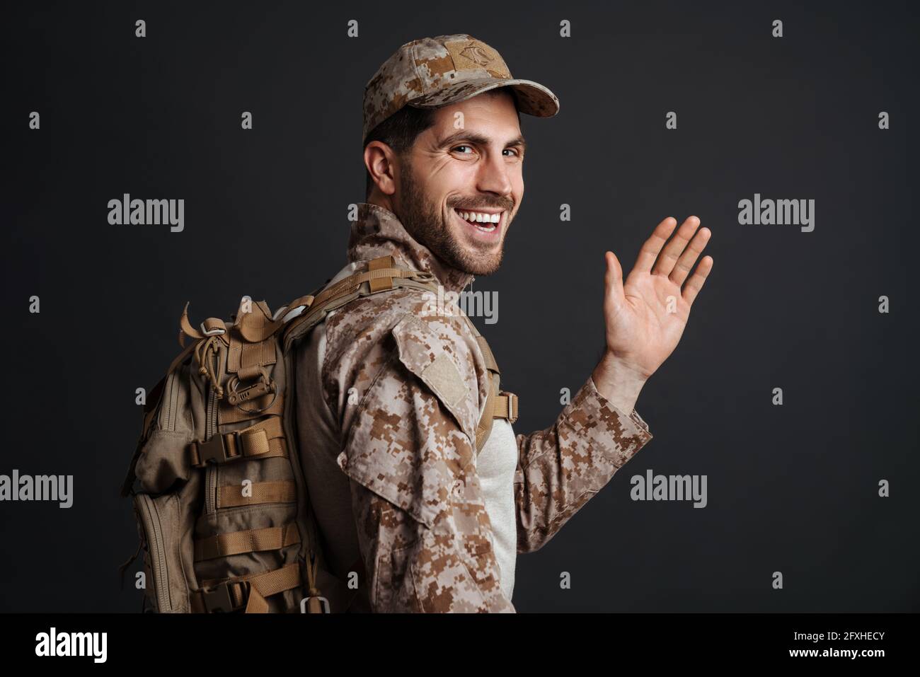 Smiling masculine military man waving hand while posing with backpack ...